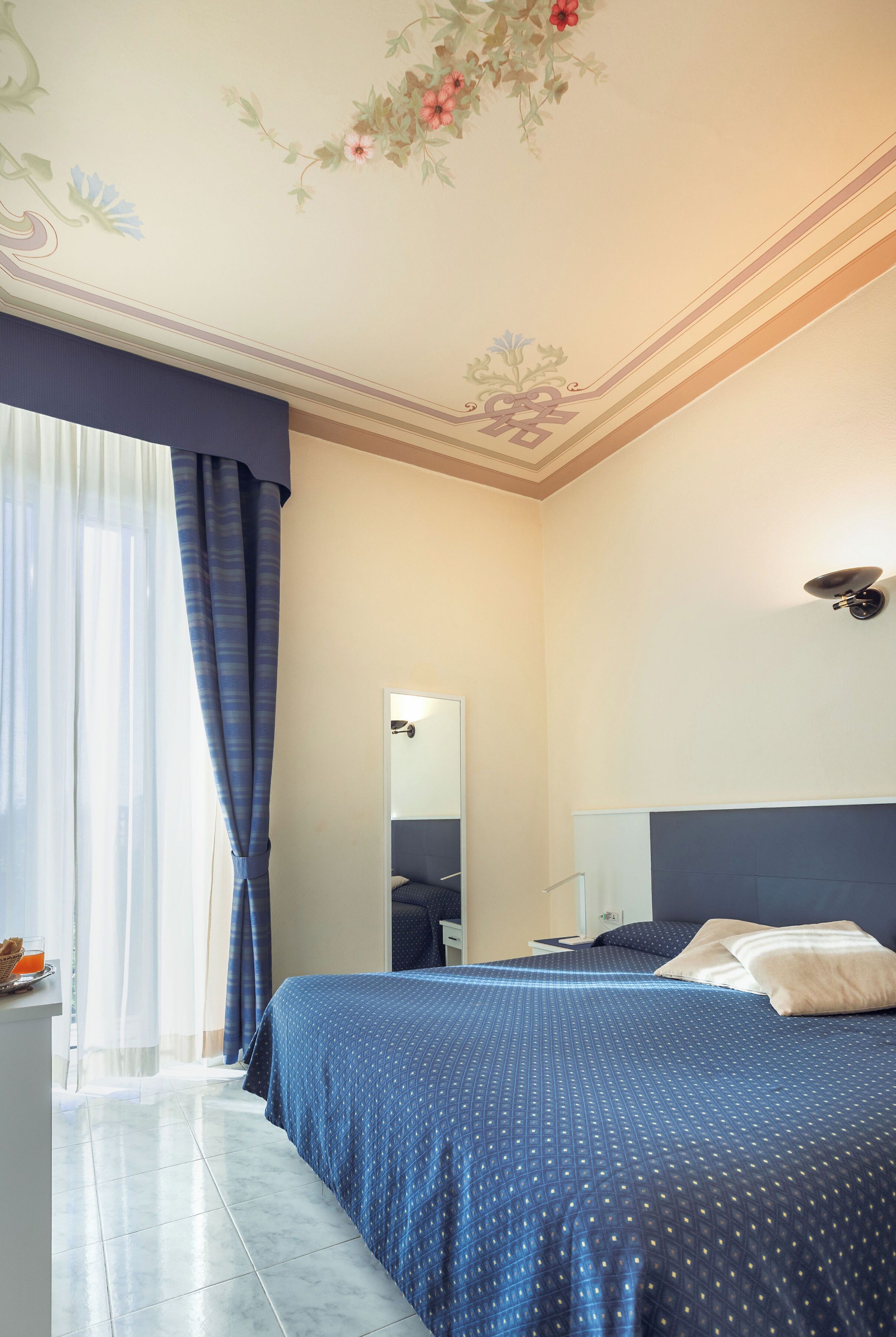 Quarto de hotel com uma cama, uma mesa, um espelho e uma janela com cortinas.