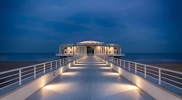Um calçadão à beira-mar que conduz a um edifício com cúpula e duas alas, apresentando uma escadaria central e um corrimão ao longo do caminho.