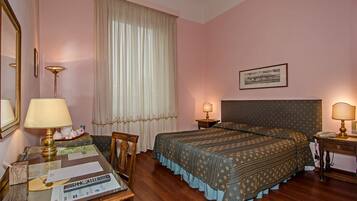 Quarto de hotel com cama, escrivaninha com luminária, cadeira, mesa de cabeceira e um quadro na parede.