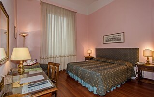 Quarto de hotel com cama, escrivaninha com luminária, cadeira, mesa de cabeceira e um quadro na parede.