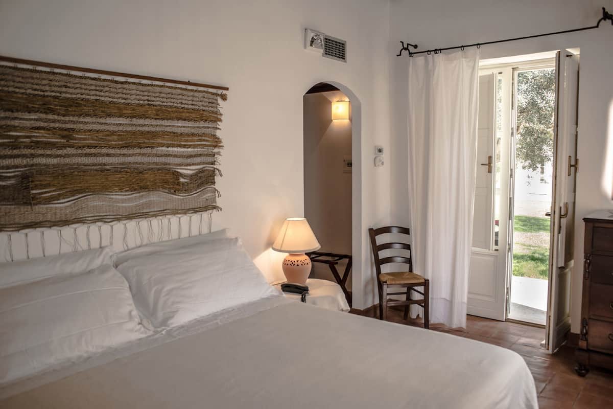 Quarto com cama, mesa de cabeceira, luminária, cadeira e porta que dá para uma varanda.