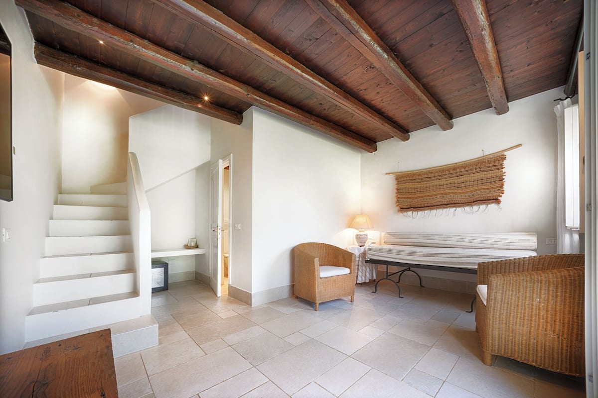 Um quarto espaçoso com teto de madeira, escada, cadeira de vime, um banco e uma cama.