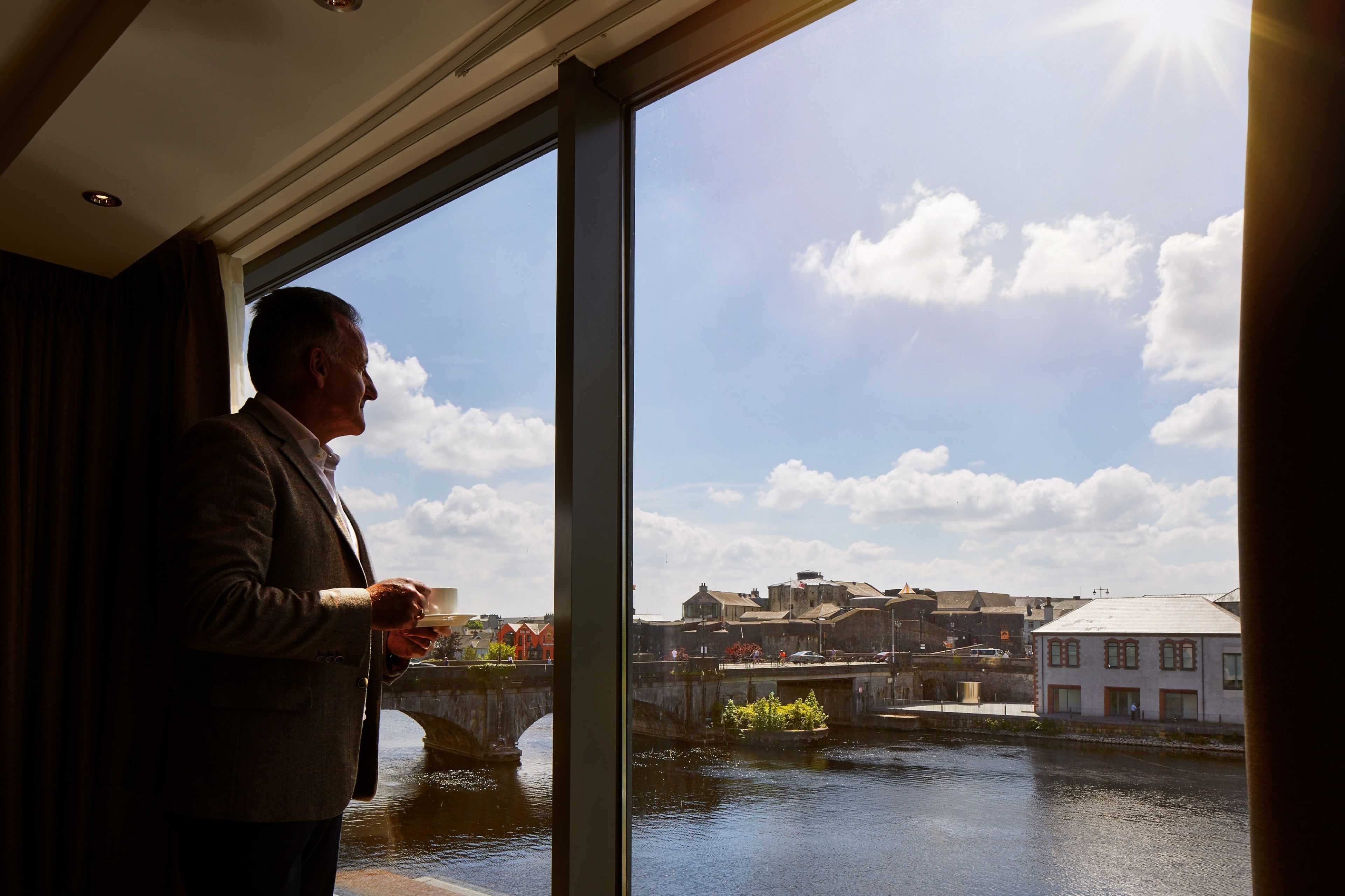 Um homem parado junto a uma janela com vista para um rio, com edifícios e uma ponte ao fundo.