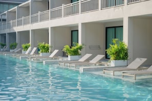 Piscina com espreguiçadeiras e plantas em vasos, rodeada por um edifício branco com varandas.