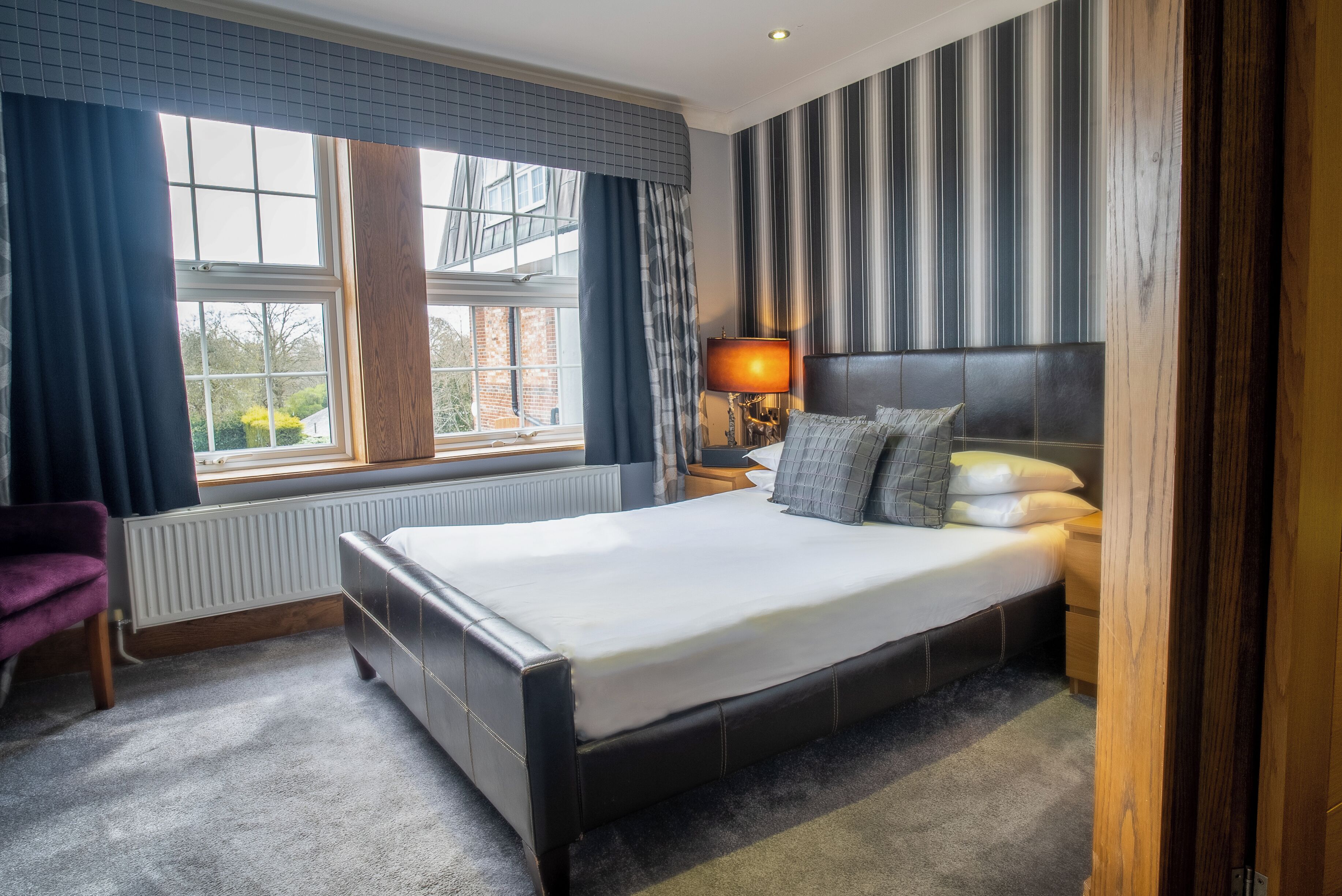 Quarto de hotel com uma cama grande, janela com cortinas, mesa de cabeceira com abajur e radiador.