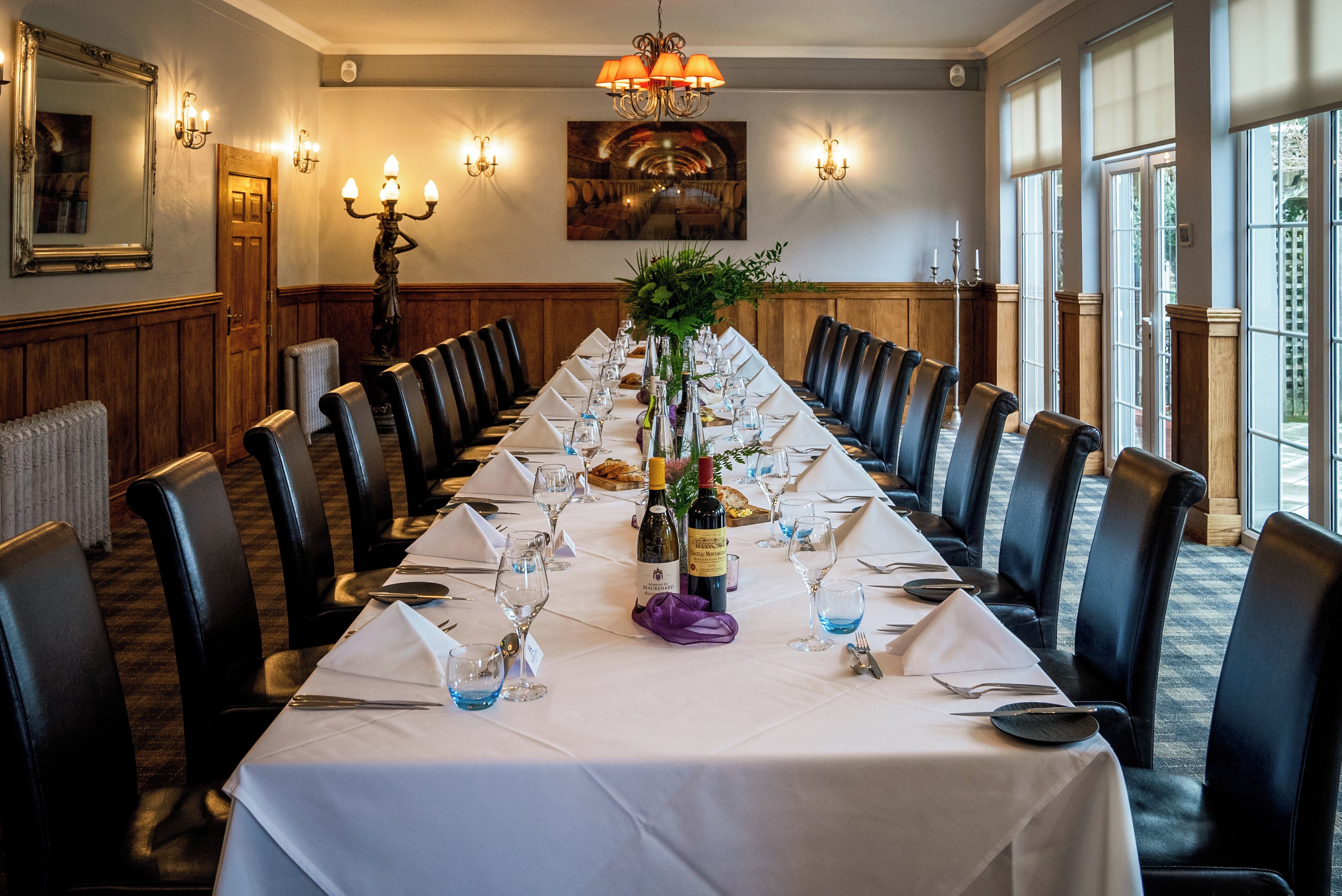 Uma longa mesa de jantar posta com cadeiras, garrafas de vinho e copos, em um ambiente com painéis de madeira e janelas amplas.