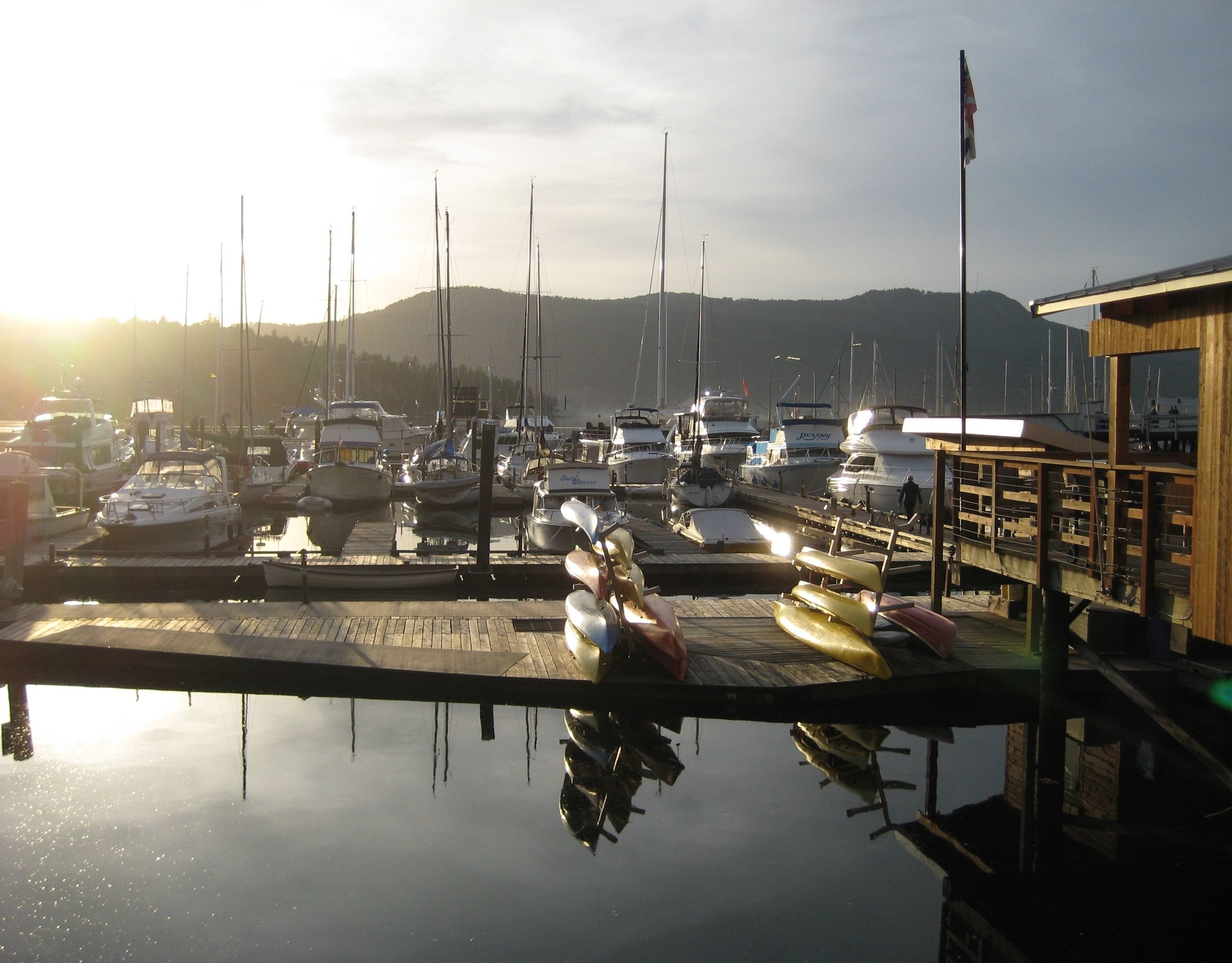 Uma marina com barcos atracados, um píer de madeira e uma montanha ao fundo.