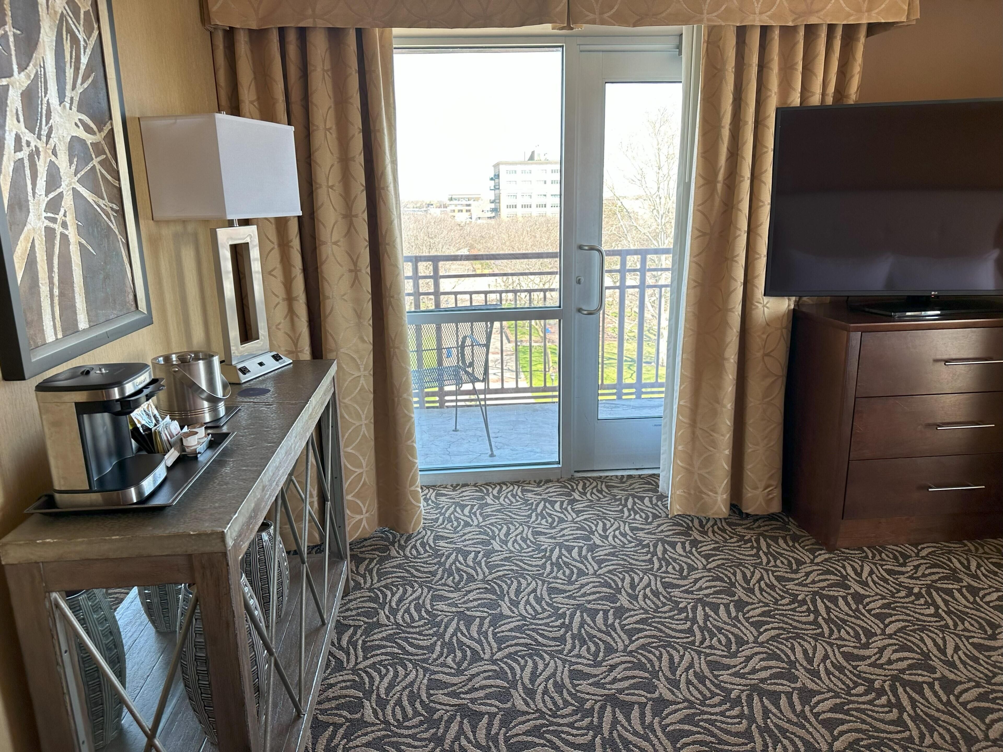 Quarto de hotel com varanda, televisão, máquina de café e carpete estampado.