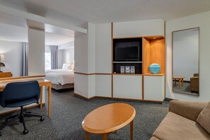 Quarto de hotel com cama, escrivaninha, cadeira, mesa pequena, televisão e espelho.