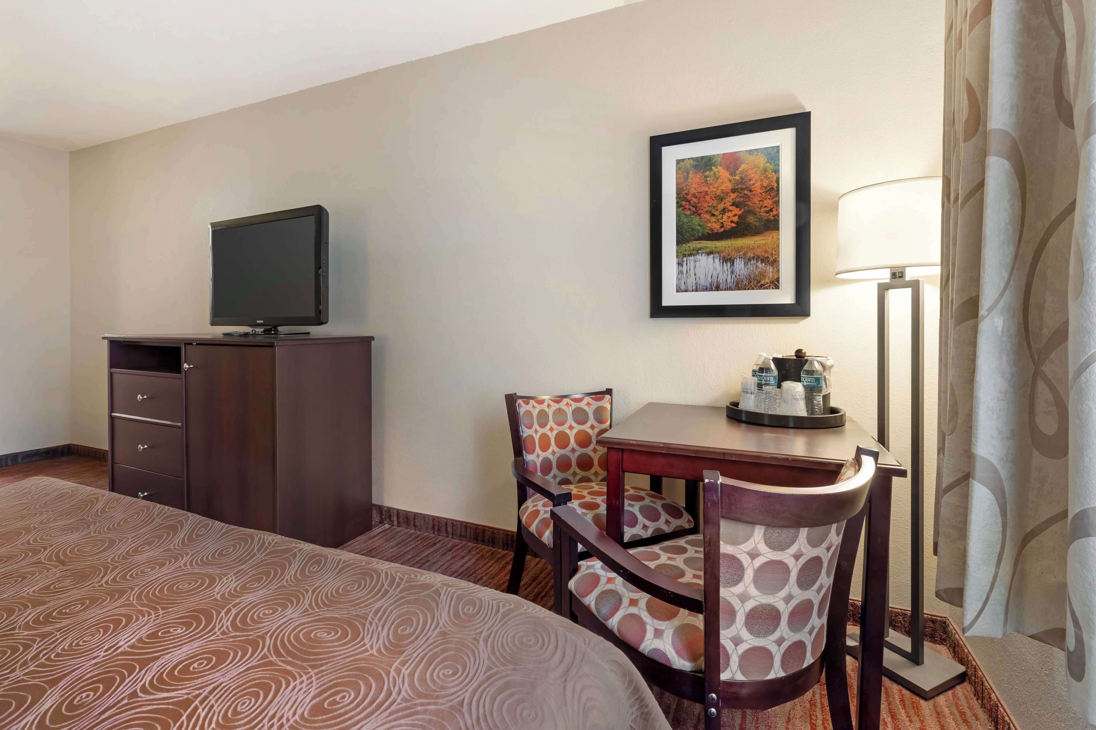 Quarto de hotel com cama, escrivaninha com cadeira, televisão, quadro emoldurado, abajur e janela com cortinas.