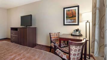 Quarto de hotel com cama, escrivaninha com cadeira, televisão, quadro emoldurado, abajur e janela com cortinas.