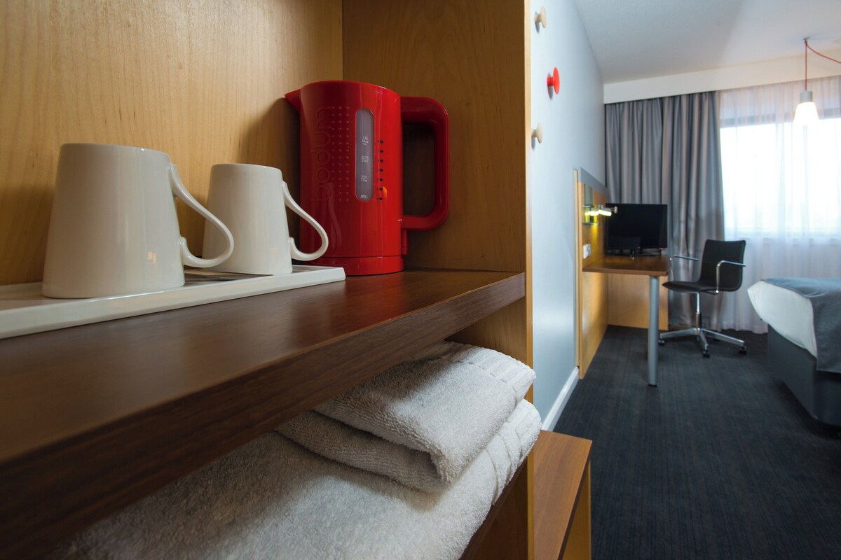 Quarto de hotel com uma estante de madeira que acomoda dois copos brancos, uma cafeteira vermelha e uma toalha dobrada.