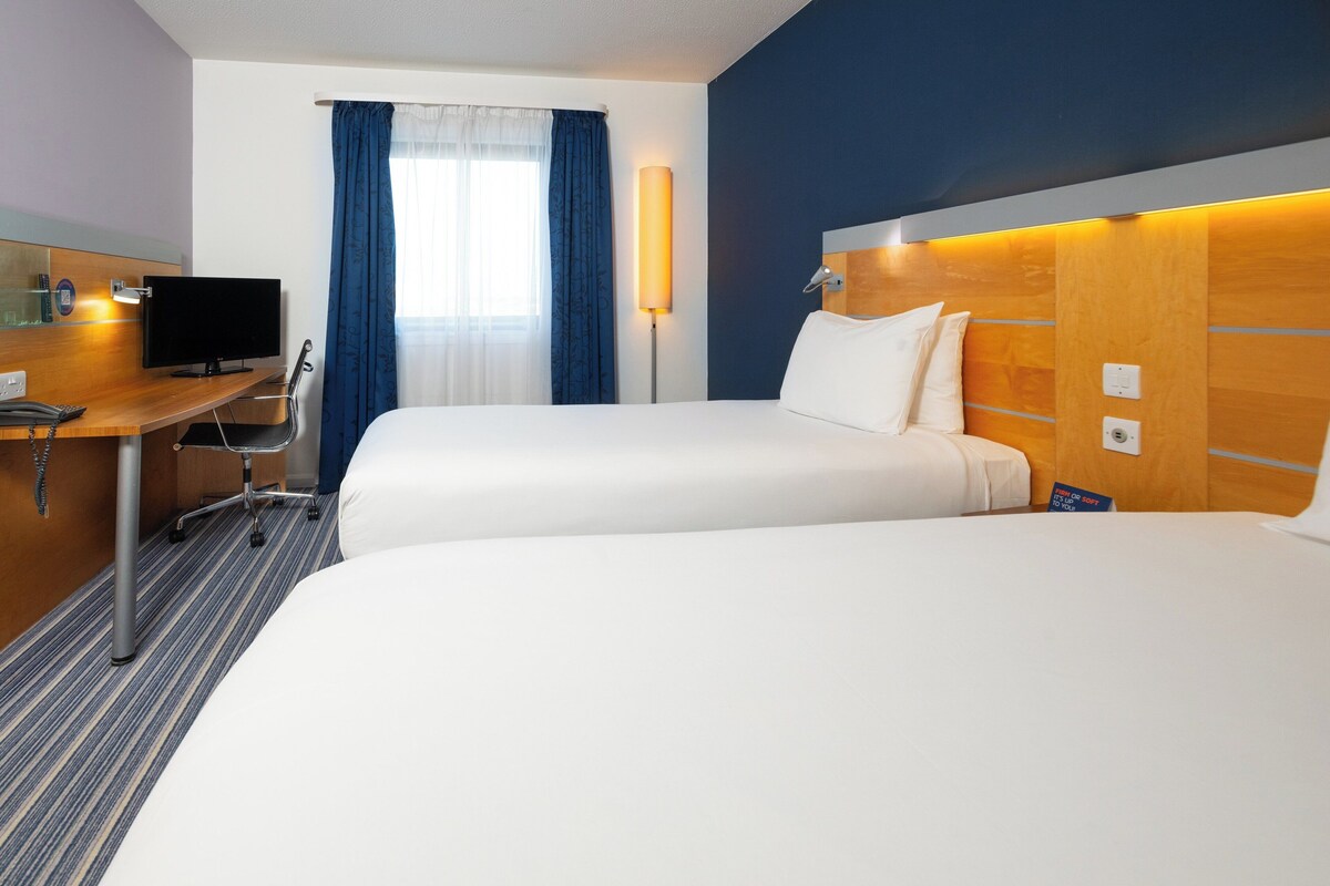 Quarto de hotel com duas camas, uma mesa de trabalho com computador, uma luminária e uma janela com cortinas.