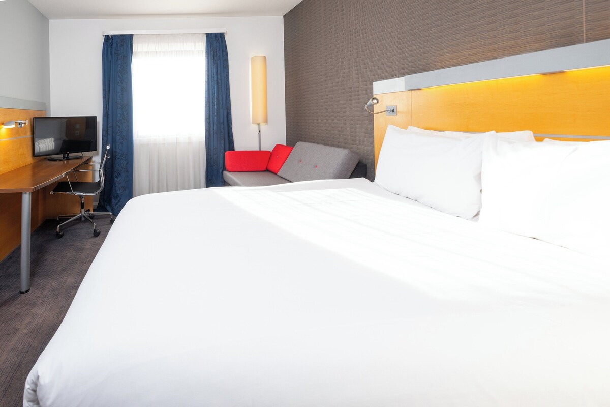 Quarto de hotel com uma cama grande, uma escrivaninha com computador, uma cadeira e uma televisão.