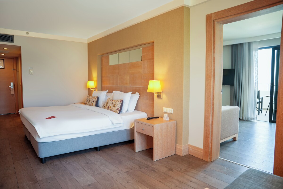 Quarto de hotel com uma cama grande, abajures, cabeceira de madeira e vista para a varanda.
