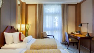 Quarto de hotel com duas camas, uma escrivaninha, uma cadeira, um abajur e uma janela grande com cortinas.