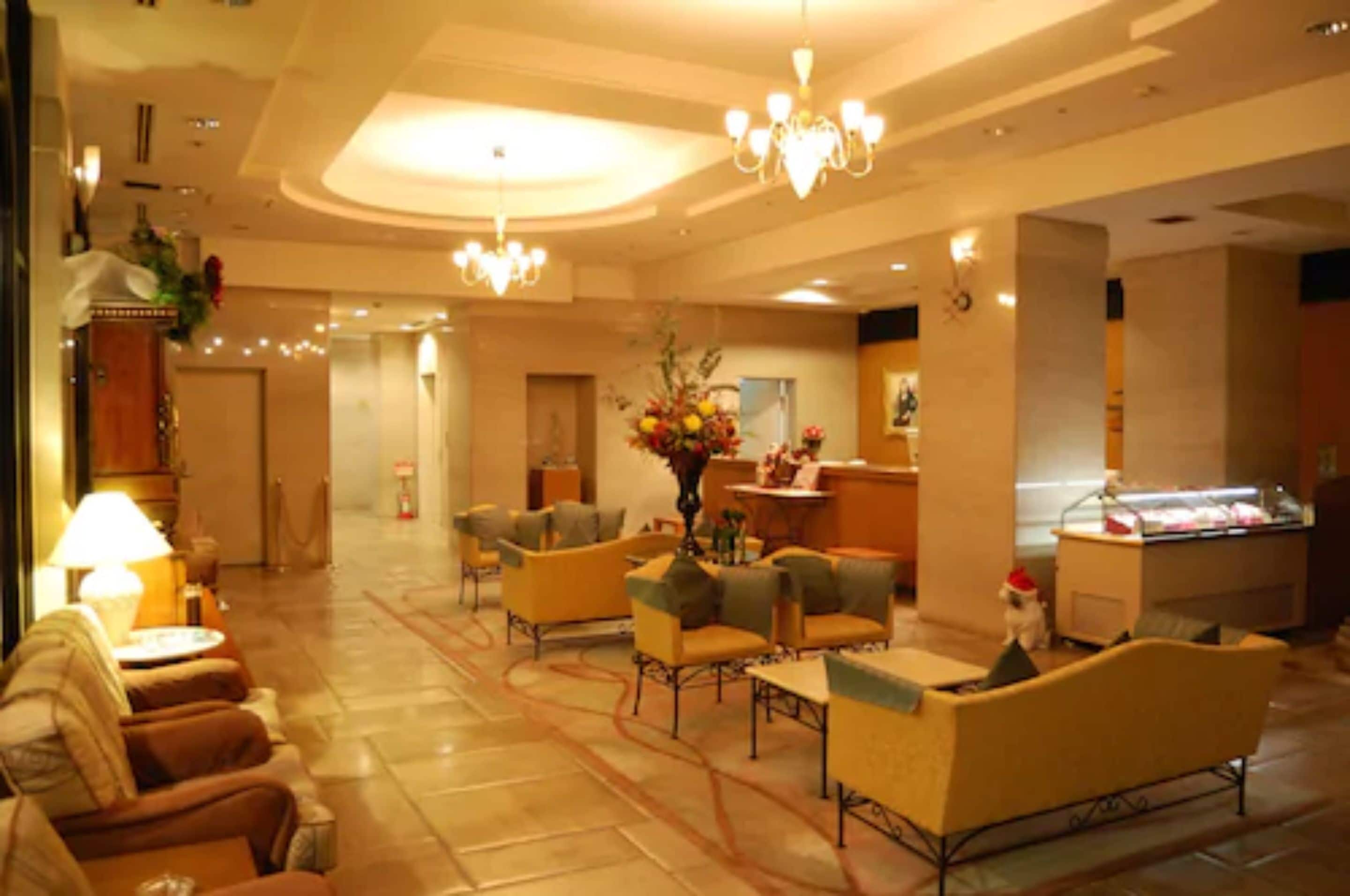 Um hall de hotel com balcão de recepção, área de estar com sofás e poltronas, um lustre e uma vitrine.
