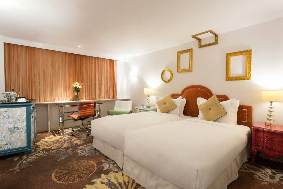 Quarto de hotel com duas camas, uma escrivaninha, uma cadeira, um armário e uma janela com cortinas.