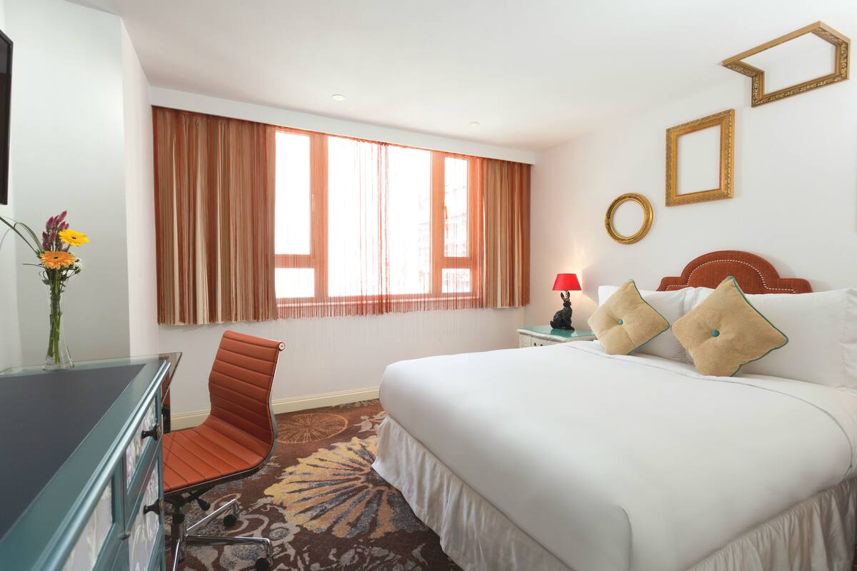 Quarto de hotel com cama, escrivaninha, cadeira, janela com cortinas e parede com quadros emoldurados.