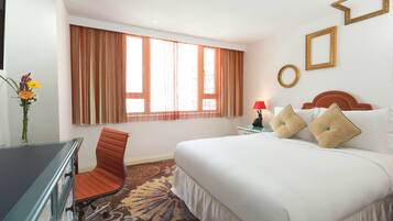 Quarto de hotel com cama, escrivaninha, cadeira, janela com cortinas e parede com quadros emoldurados.