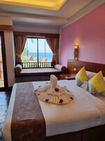 Quarto de hotel com uma cama, mesa de cabeceira, janela com cortinas e vista para o oceano.