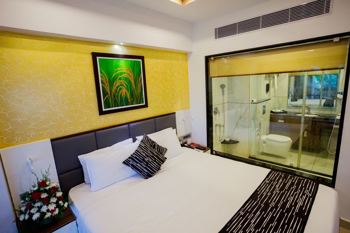 Quarto de hotel com uma cama, uma mesa de cabeceira com um vaso de flores, um quadro na parede e um banheiro com divisória de vidro.