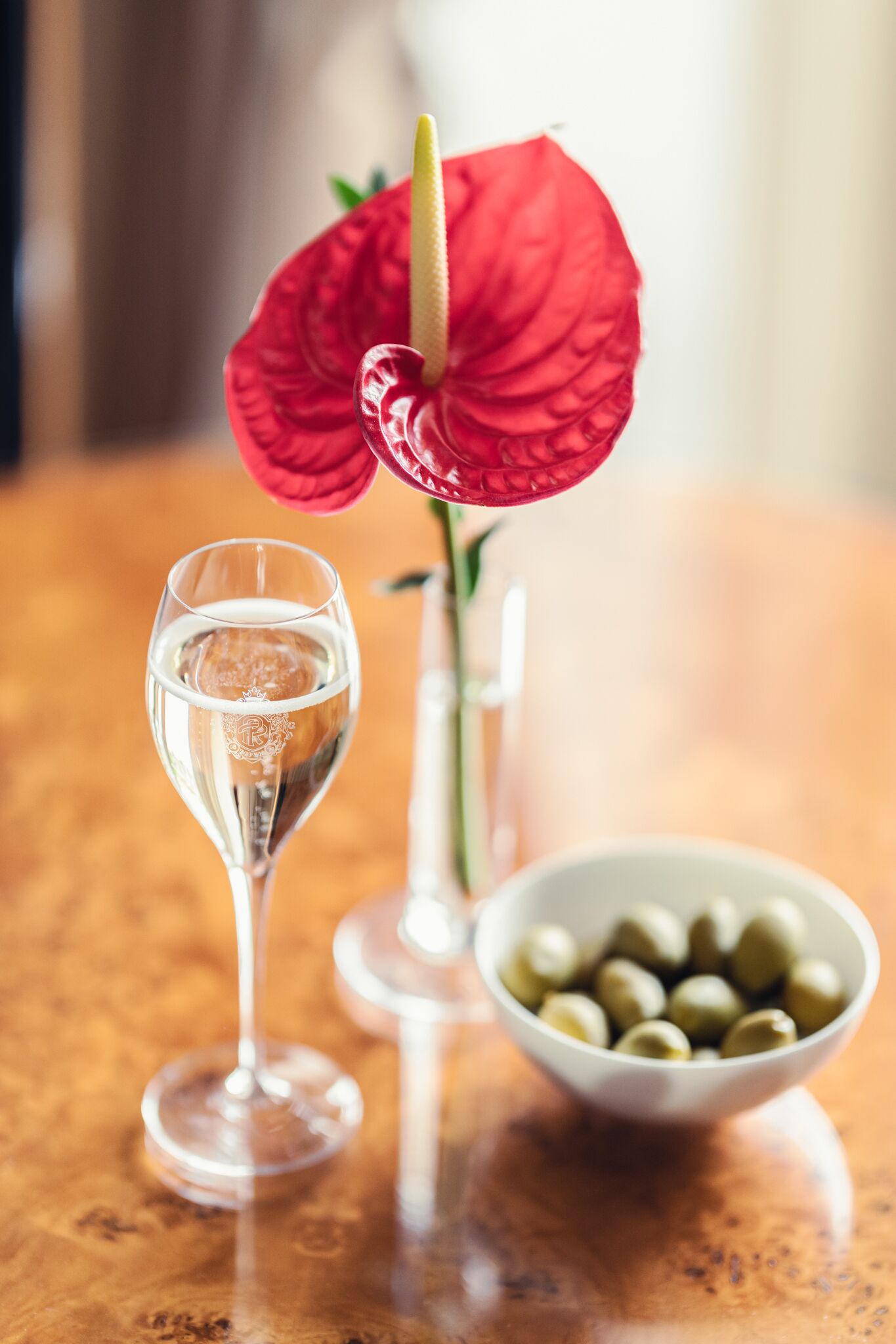 Uma taça de champanhe cristalina, uma travessa de azeitonas e uma flor vermelha sobre uma mesa de madeira.