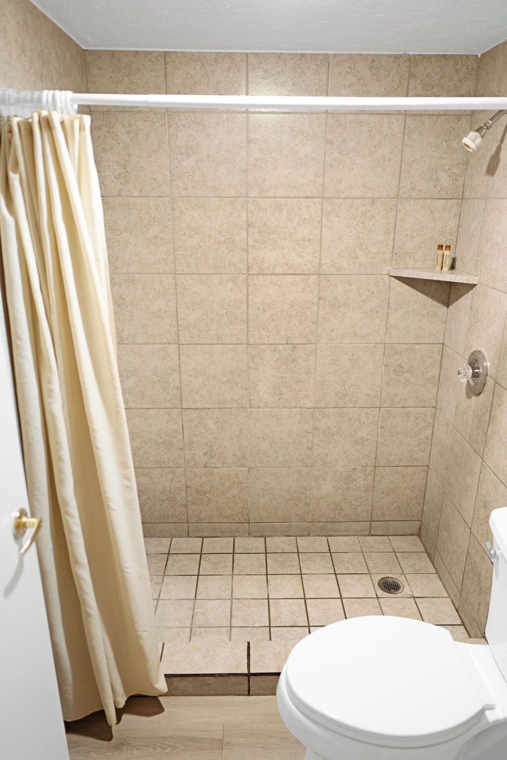 Casa de banho com azulejos bege, chuveiro, sanita e uma prateleira com produtos de higiene pessoal.