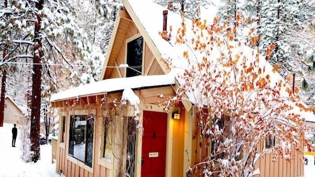 Uma cabana de madeira com porta vermelha, rodeada por árvores cobertas de neve e um arbusto sem folhas.