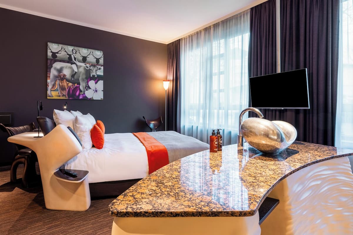 Quarto de hotel com cama, escrivaninha com pia em formato de coração, um quadro na parede e televisão.