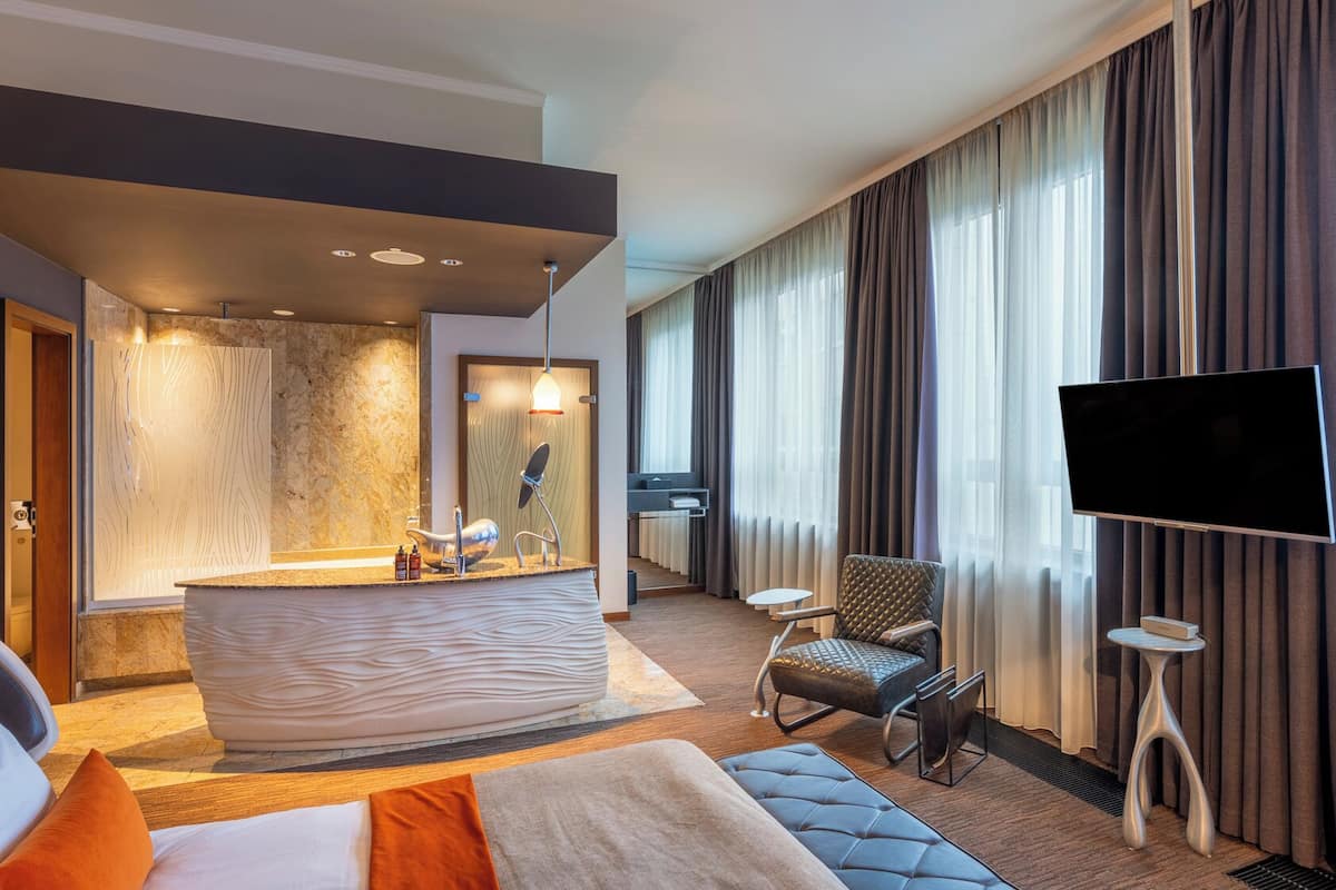 Quarto de hotel moderno com uma cama grande, área de estar com poltrona, televisão de tela plana, banheiro com banheira e uma grande janela com cortinas.