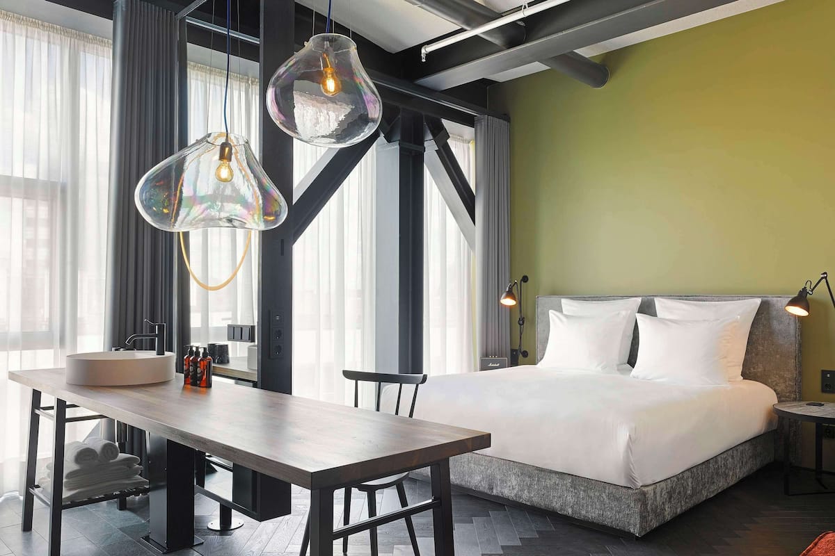 Quarto de hotel moderno com uma cama grande, uma escrivaninha de madeira e duas luminárias pendentes.
