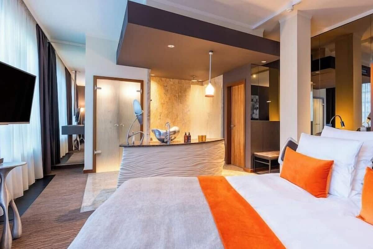 Quarto de hotel com cama, televisão, banheiro com espelho e escrivaninha.