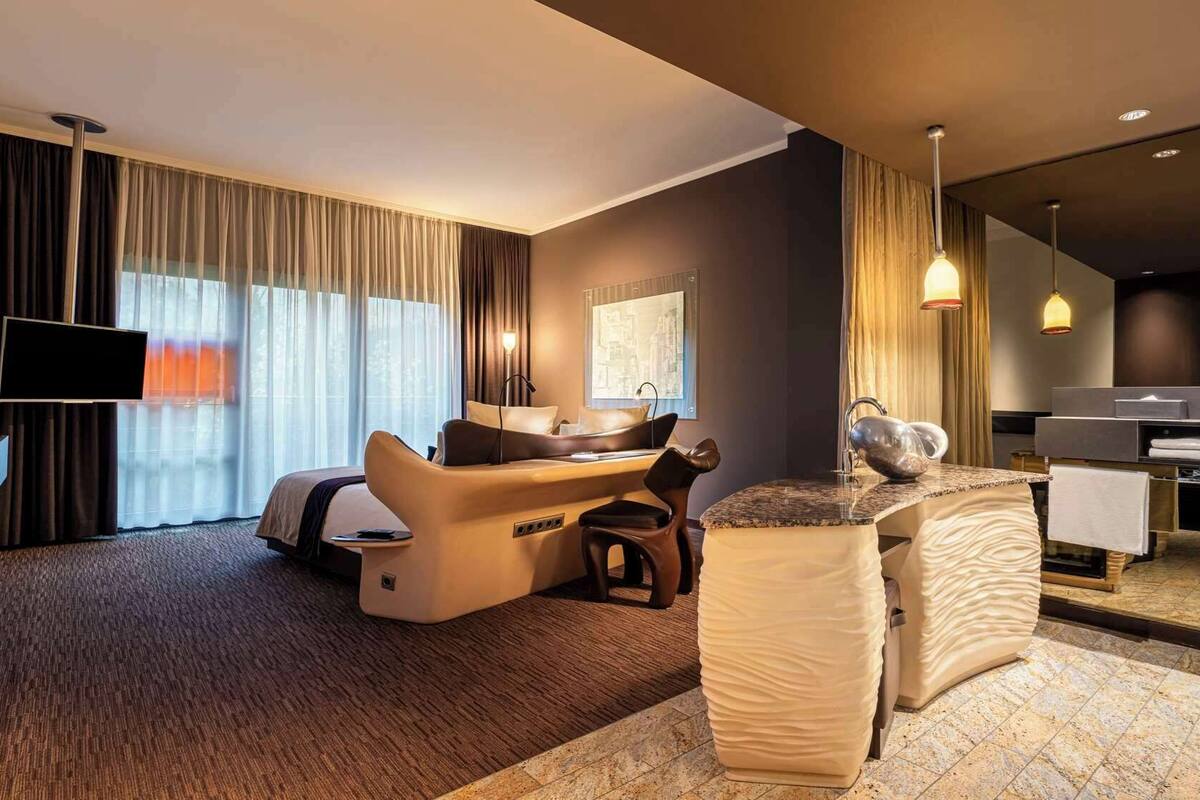 Quarto de hotel moderno com sofá-cama, escrivaninha, televisão e banheiro com espelho e pia.