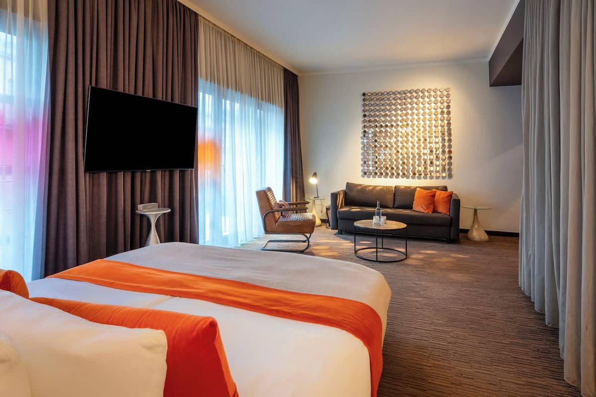 Quarto de hotel com uma cama grande, um sofá, uma poltrona, uma mesa de centro e uma televisão de tela plana.