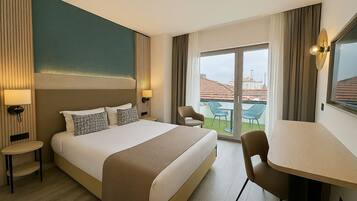 Quarto de hotel com cama, escrivaninha, cadeira, mesinha e vista para o exterior.