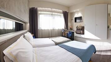 Quarto de hotel com duas camas, uma escrivaninha, uma cadeira, um guarda-roupa e uma janela grande com cortinas.
