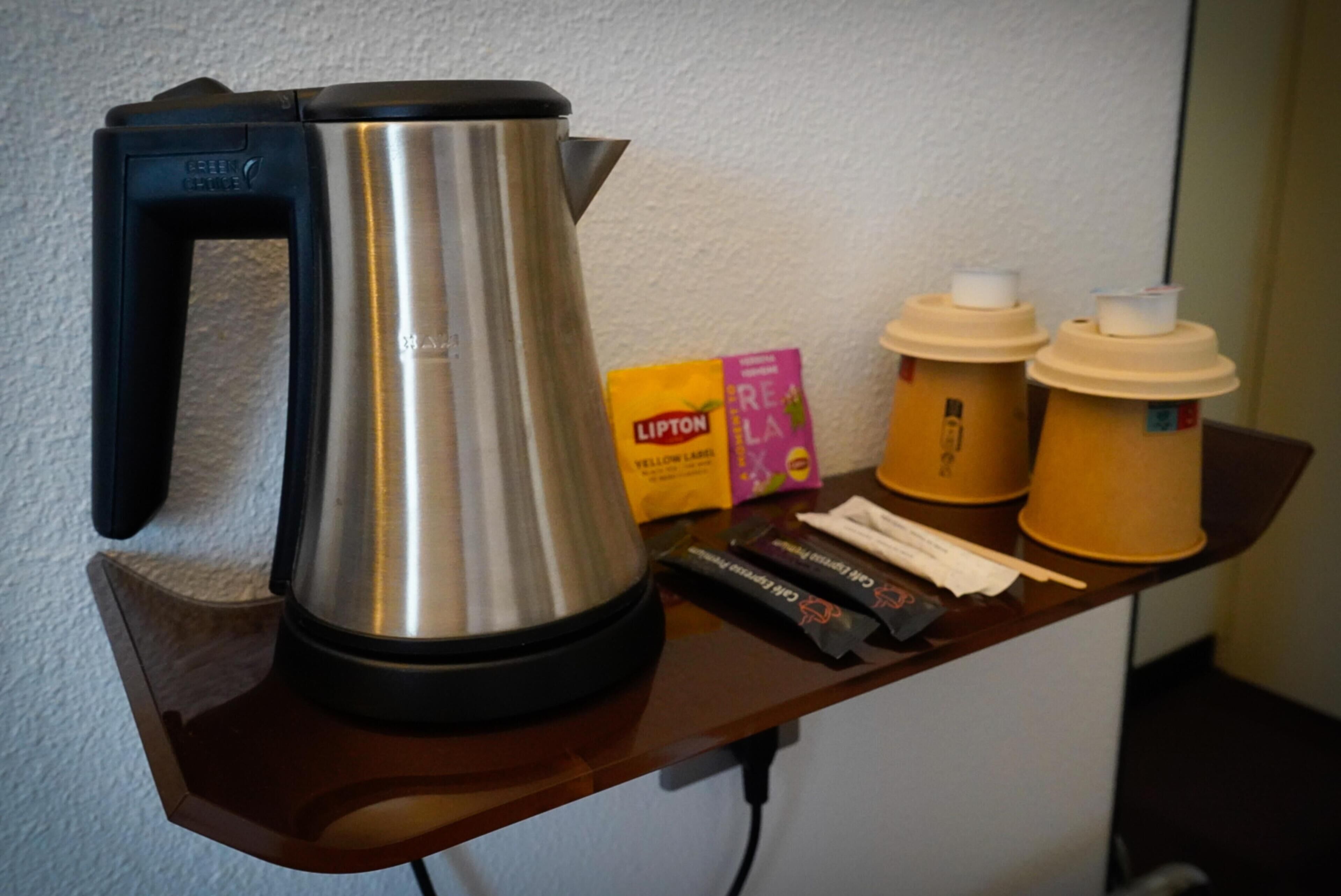 Uma chaleira de aço inoxidável sobre uma prateleira de madeira, com saquinhos de chá Lipton, uma caixa de chá Lipton e duas chávenas de café descartáveis.