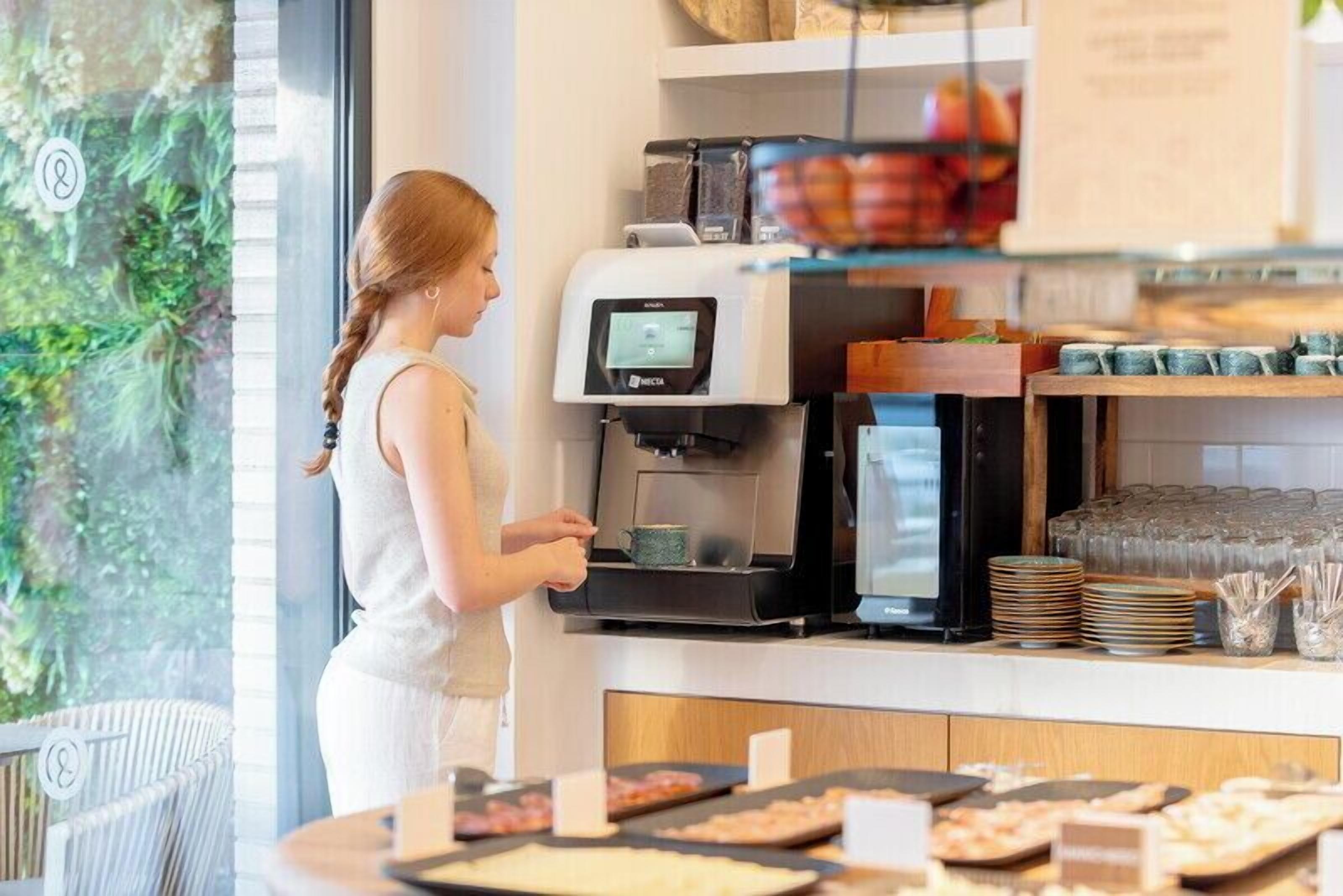 Máquina de café, balcão com alimentos e janela com vegetação.