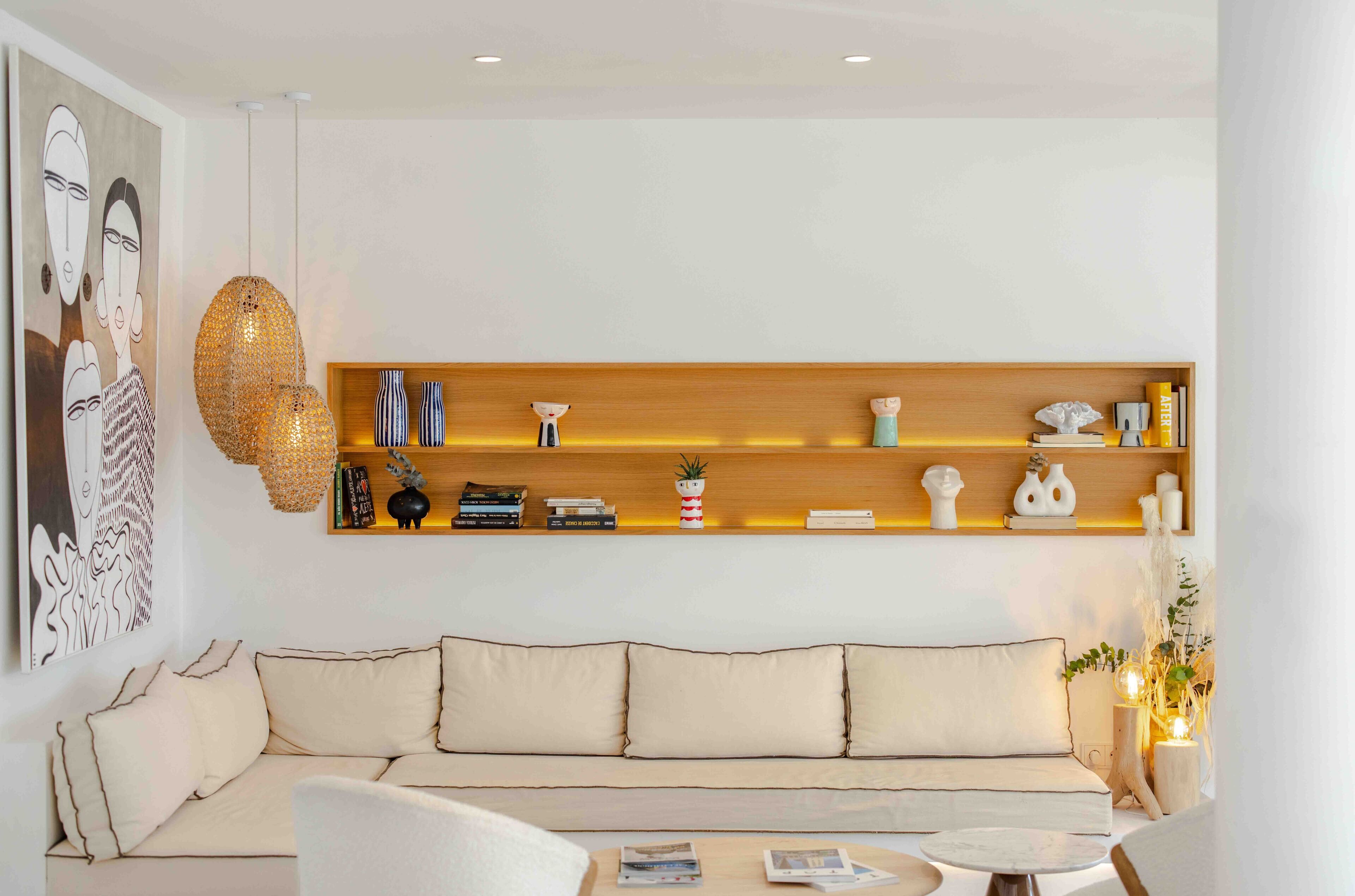 Uma sala de estar moderna com um sofá bege, uma estante de madeira com livros e objetos decorativos, e um grande quadro abstrato na parede.