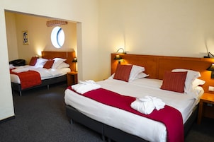 Quarto de hotel com duas camas, cada uma com cabeceira de madeira, roupa de cama branca e almofadas decorativas vermelhas. Há luminárias laterais e uma pequena mesa com abajur.