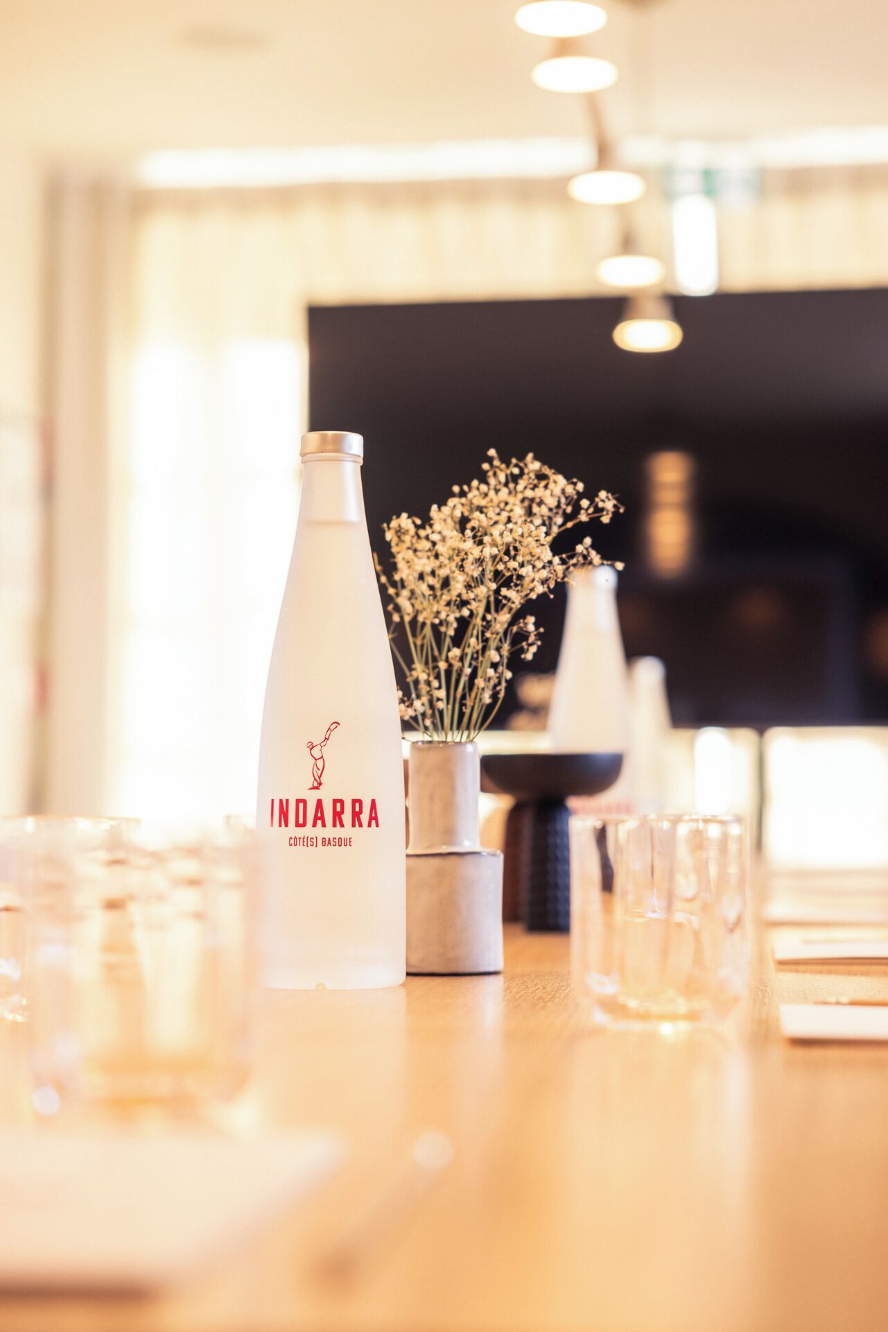 Uma garrafa de vinho Indarra Cote(s) Basque sobre uma mesa, com um vaso de flores e vários copos.