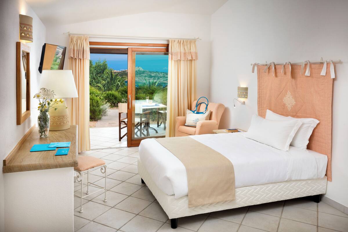 Quarto de hotel com cama, escrivaninha com luminária, cadeira e vista para o exterior.