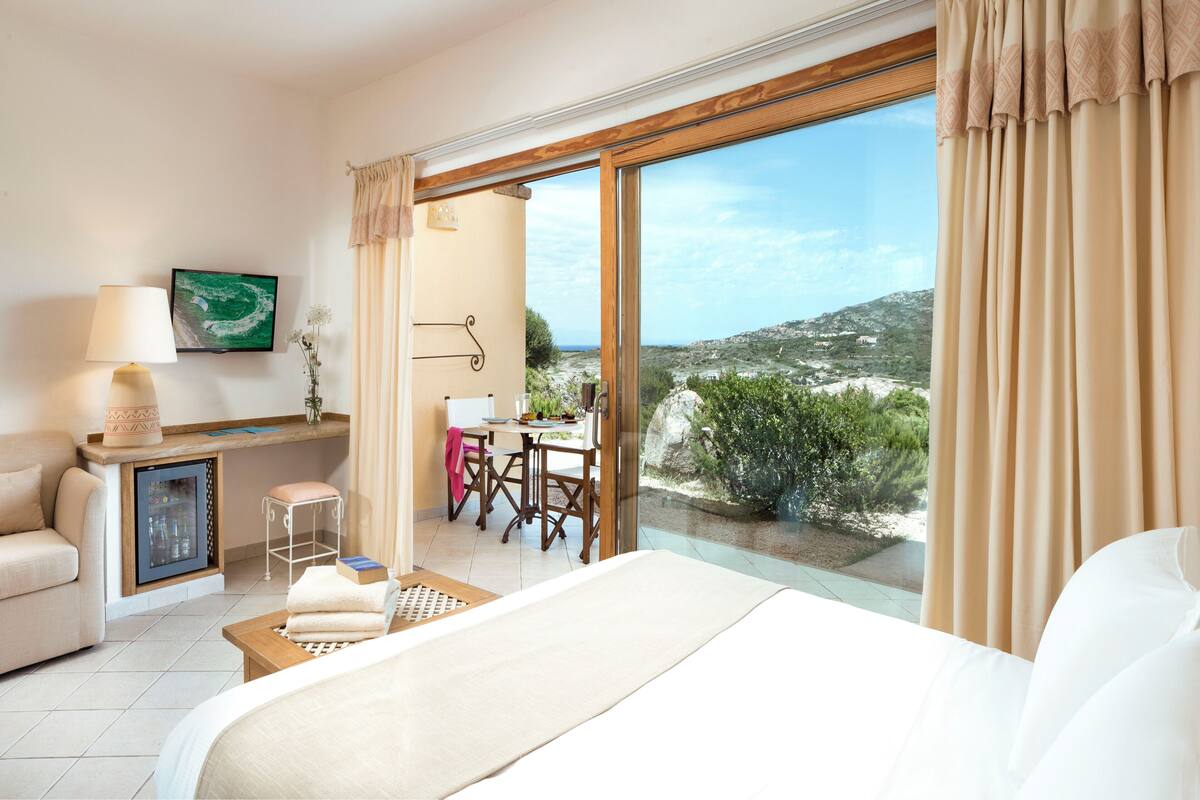 Quarto de hotel com cama, escrivaninha, cadeira, mesa pequena, sofá, TV e uma grande janela com vista para a paisagem.