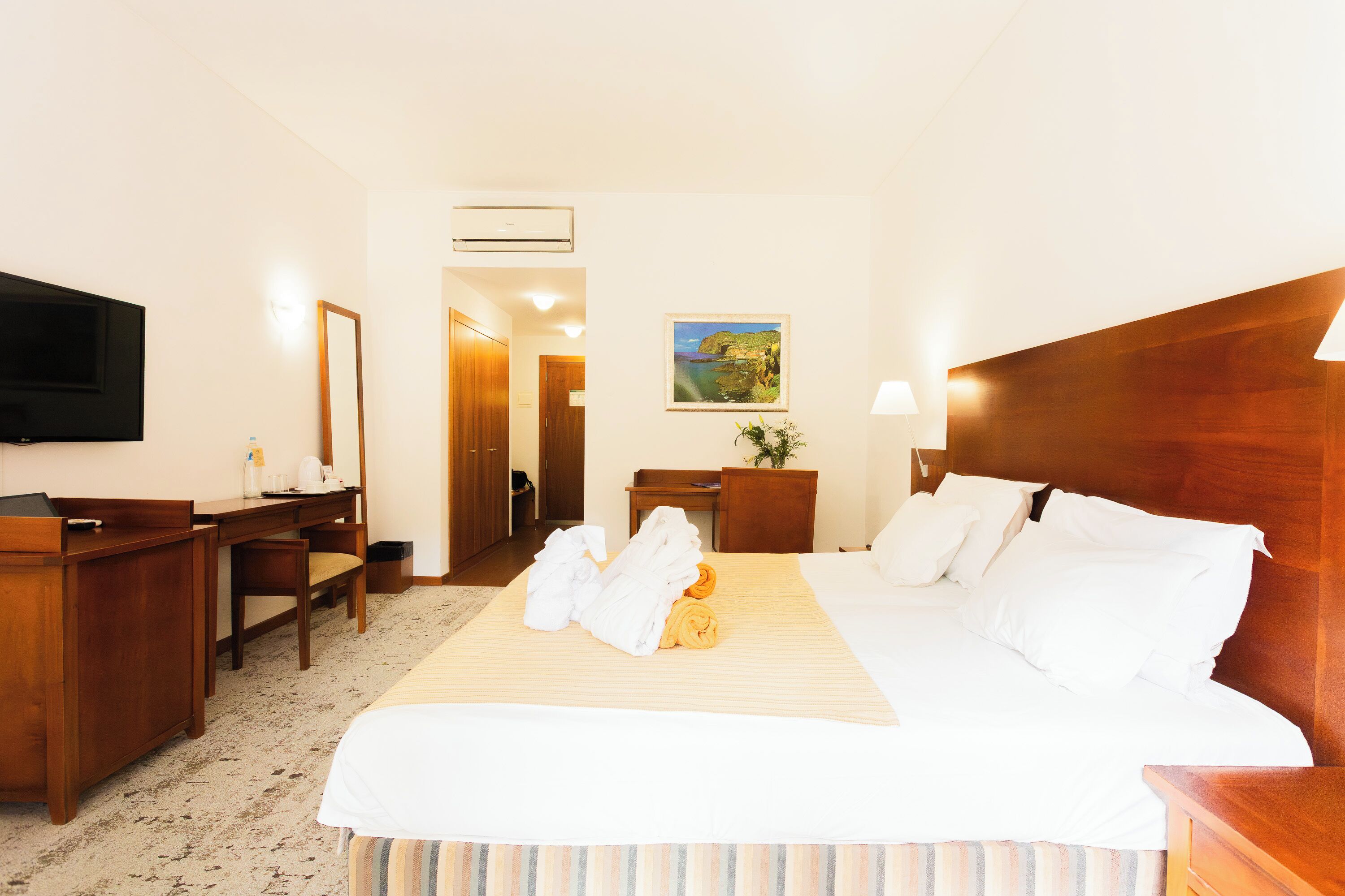Quarto de hotel com cama, escrivaninha, cadeira, televisão e um quadro na parede.