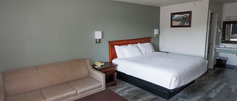 Quarto de hotel com cama, sofá, mesa pequena, mesa de cabeceira, luminária na parede e um quadro.