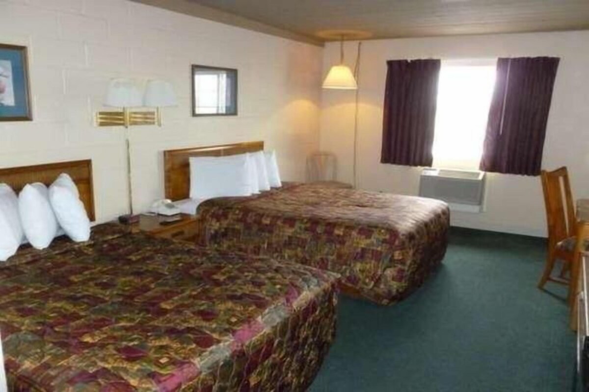 Quarto de hotel com duas camas, uma escrivaninha, uma cadeira, um abajur e uma janela com cortinas.