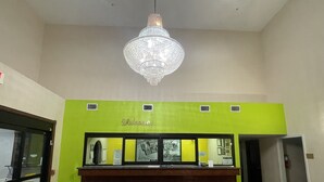 Uma área de recepção com parede verde, um letreiro com a palavra 'Welcome!' e um balcão com vitrine de vidro, além de um candelabro.