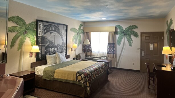 Quarto de hotel com uma cama, uma escrivaninha, uma cadeira, um abajur e uma grande pintura de uma palmeira e uma ponte.