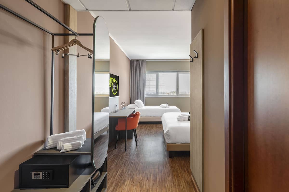 Quarto de hotel com cama, escrivaninha, cadeira, espelho, guarda-roupa e janela.